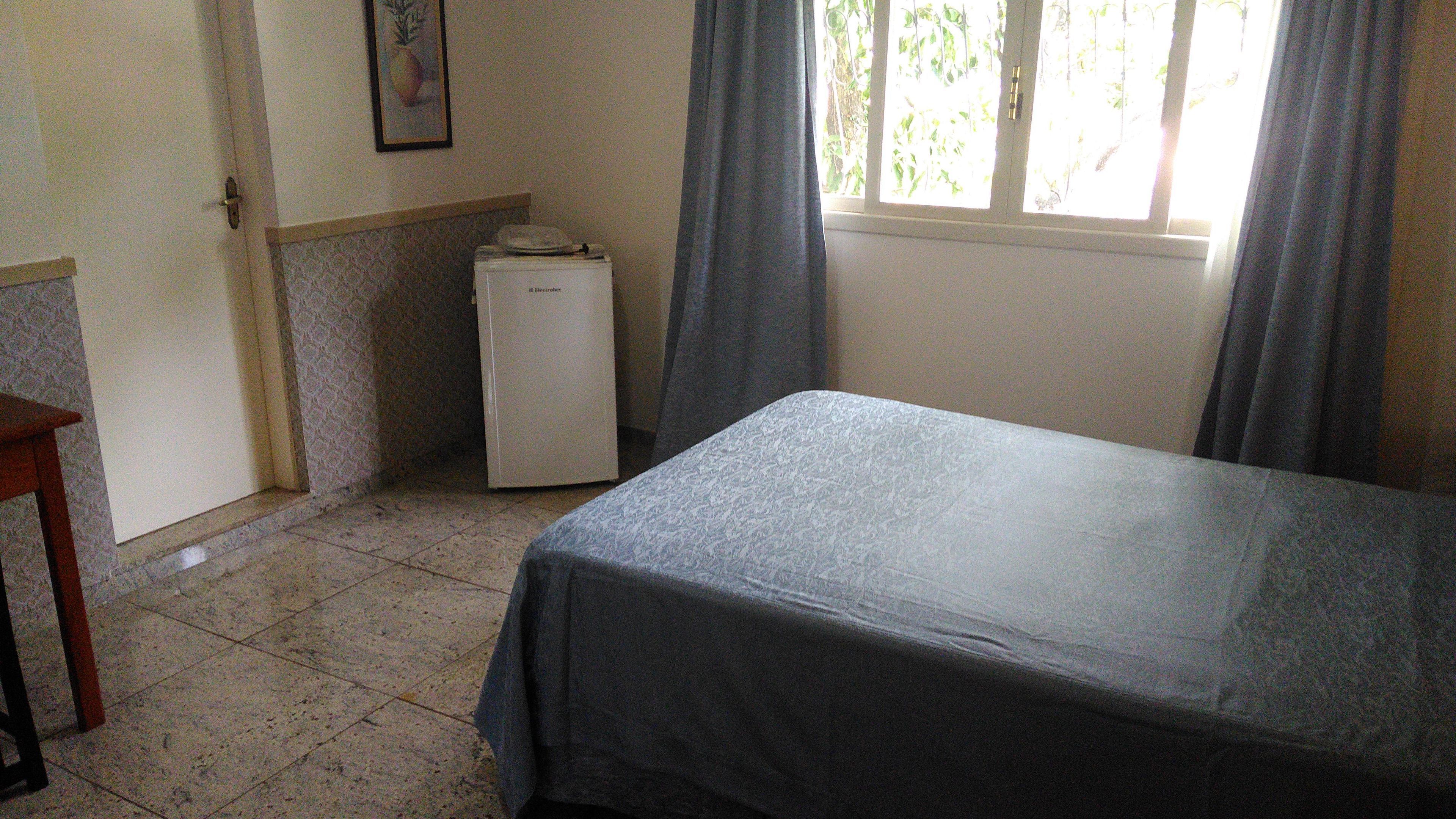 Quarto pequeno com cama, escrivaninha e geladeira.