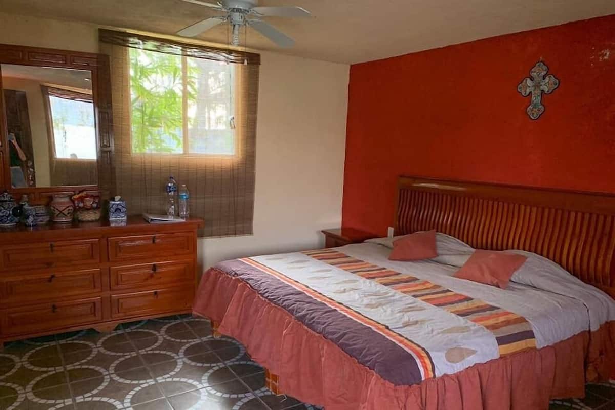 Quarto com cômoda de madeira, cama com colcha estampada, janela com persianas e ventilador de teto.