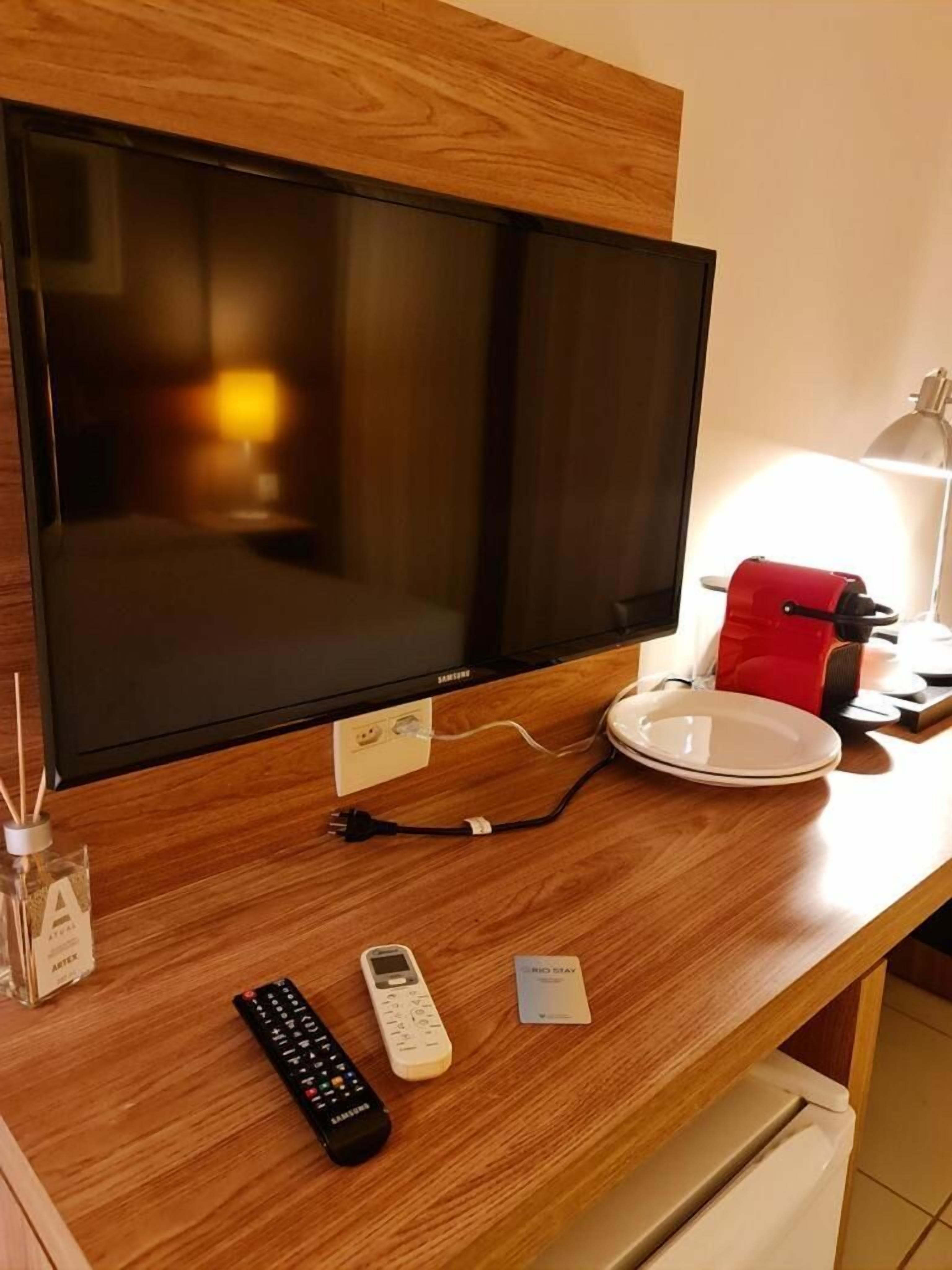 Um balcão de madeira com uma TV de tela plana, uma máquina de café vermelha, um prato branco e dois controles remotos.