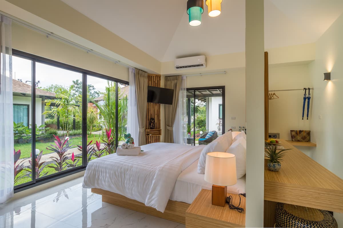 Quarto de hotel moderno com uma cama grande, mesa de cabeceira com abajur, TV na parede e vista para um jardim através de grandes janelas.