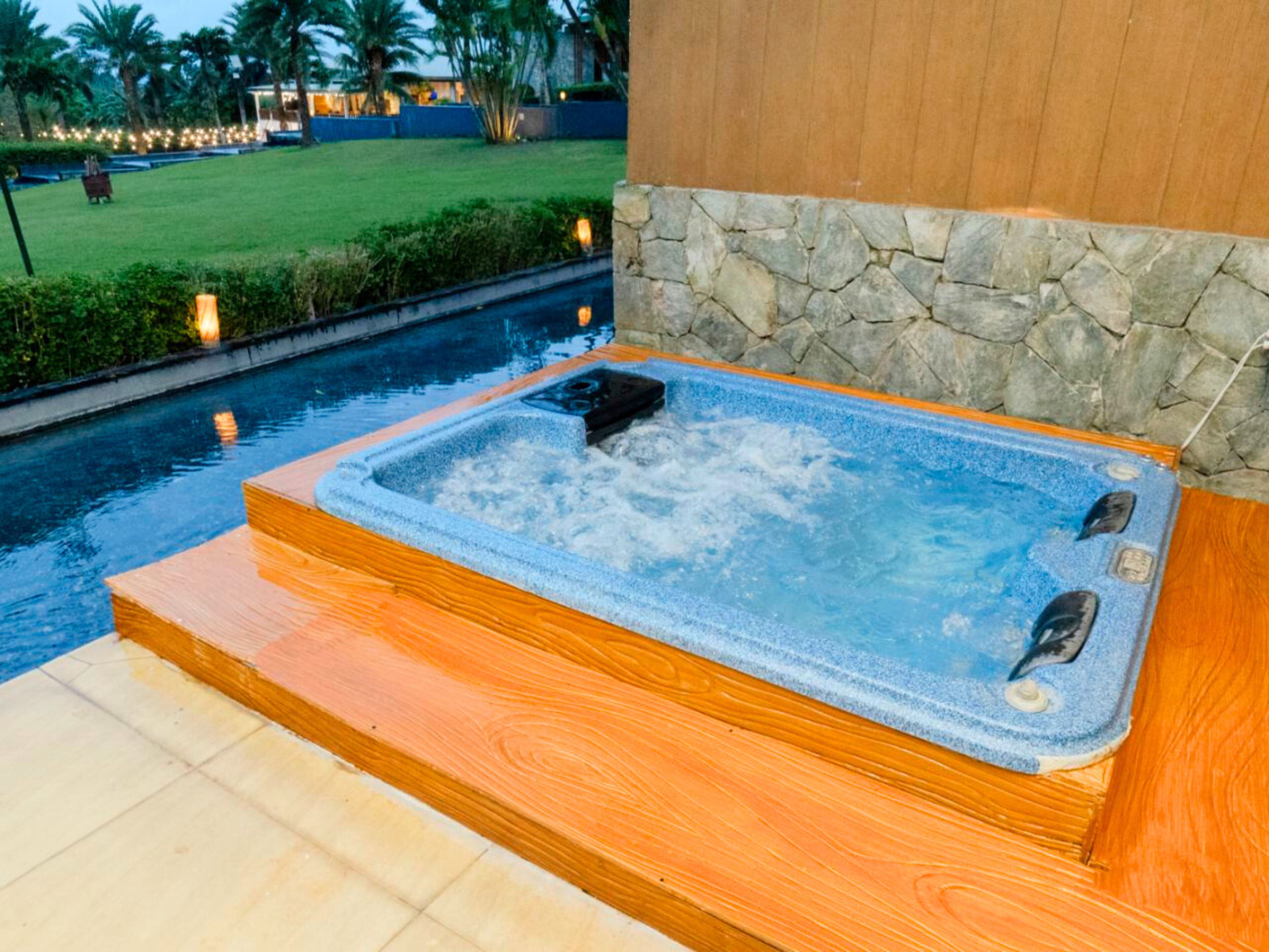 Uma banheira de hidromassagem com parede de pedra e degraus de madeira, cercada por uma piscina e vegetação.