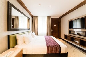 Quarto de hotel com uma cama grande, televisão, espelho e um aparador de madeira.