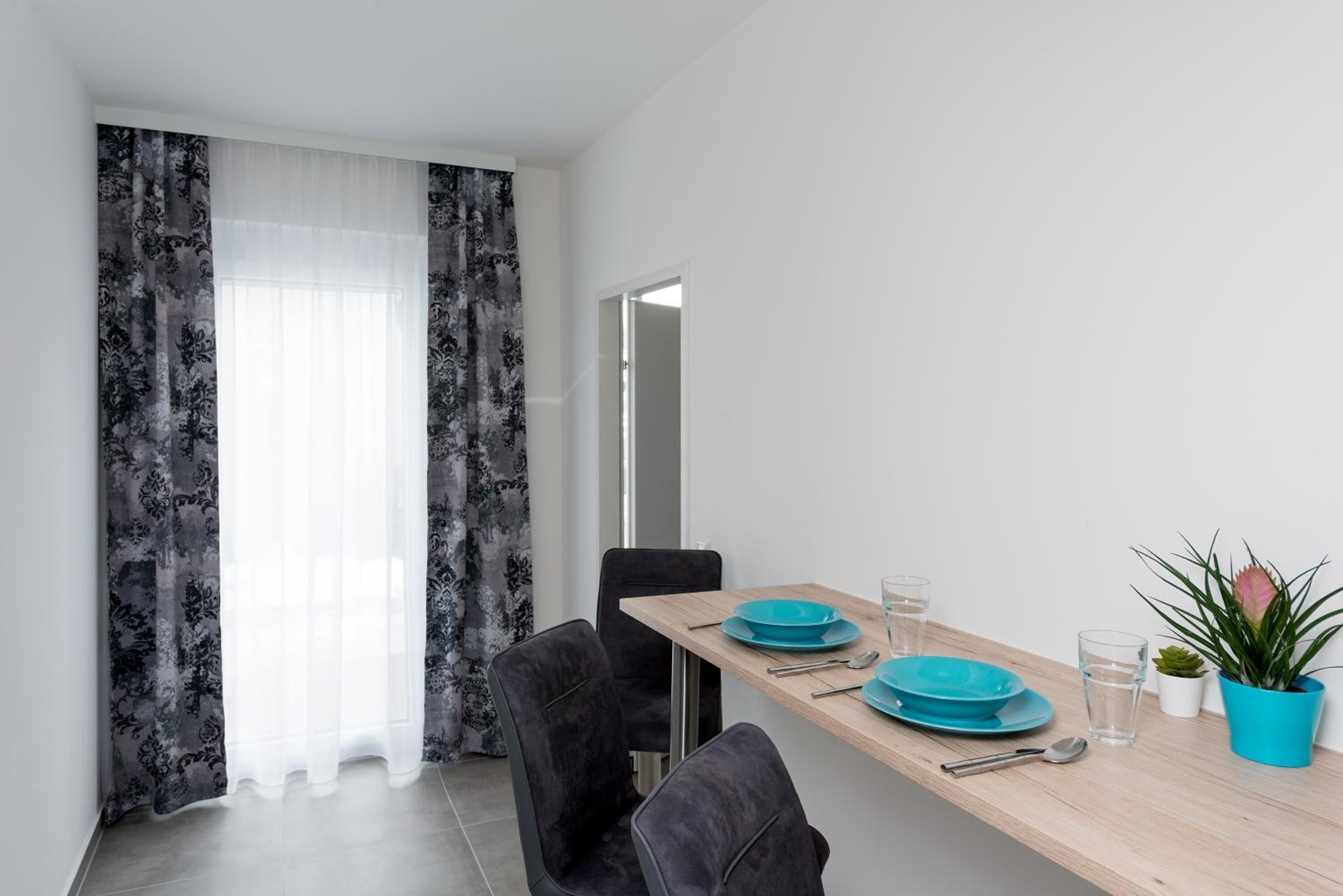 Uma área de refeições com uma mesa posta para dois, com pratos e copos azuis, e um vaso de planta à direita.