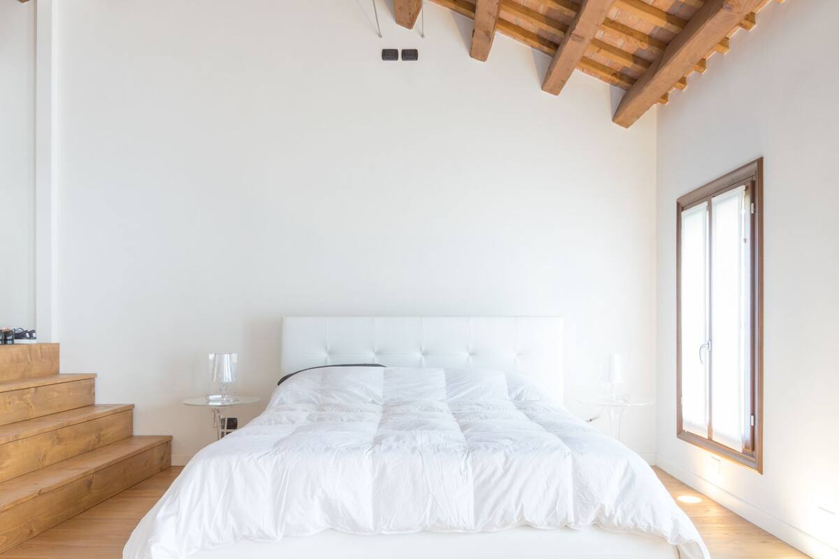 Quarto moderno com uma cama grande, escada de madeira e uma janela ampla.