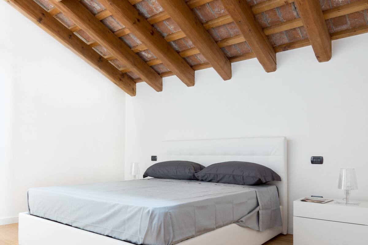 Quarto moderno com teto de madeira, uma cama branca e uma mesa de cabeceira com abajur.
