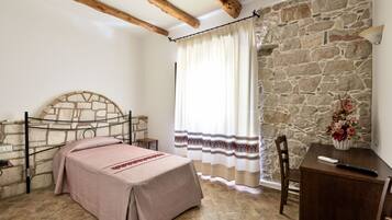 Quarto com parede de pedra, cama com colcha rosa, cabeceira de madeira, lareira de pedra e escrivaninha com um vaso de flores.