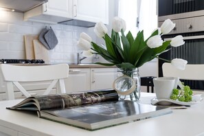 Uma cozinha com armários brancos, uma cadeira branca, uma mesa com um livro, um vaso com tulipas brancas e uma caneca branca.