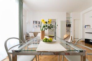 Uma área de refeições moderna com mesa de vidro, cadeiras brancas, um vaso com flores amarelas e uma tigela de maçãs verdes.