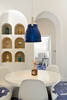 Uma sala de jantar moderna com uma mesa redonda branca e cadeiras, uma luminária pendente azul e prateleiras com objetos decorativos.