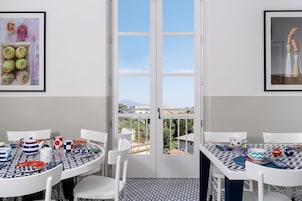 Uma área de refeições com mesas e cadeiras brancas, piso estampado e vista através de uma porta de vidro.