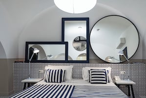 Quarto de hotel moderno com uma cama grande, duas mesas de cabeceira, um espelho redondo e um papel de parede estampado.