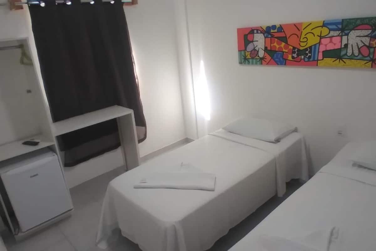 Quarto de hotel com duas camas de solteiro, uma pequena escrivaninha com cadeira, um guarda-roupa e um quadro abstrato colorido na parede.