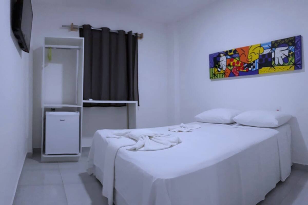 Quarto de hotel com cama, mesinha de cabeceira, guarda-roupa e um quadro abstrato colorido na parede.