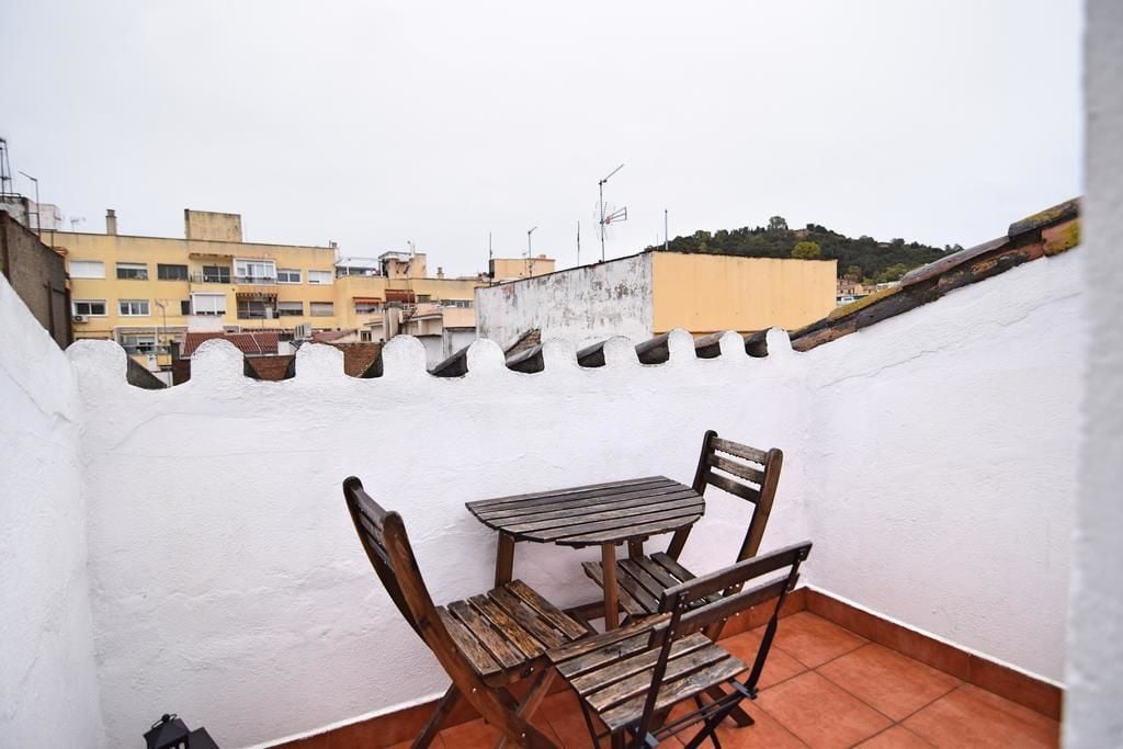 Uma pequena varanda com mesa e cadeiras de madeira, paredes brancas e vista para prédios ao fundo.