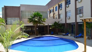 Piscina rodeada por palmeiras e espreguiçadeiras, com um edifício ao fundo.