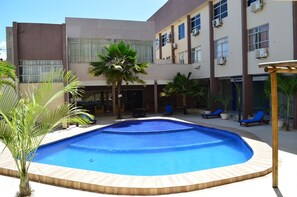 Piscina rodeada por palmeiras e espreguiçadeiras, com um edifício ao fundo.