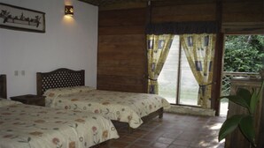 Quarto com duas camas, paredes de madeira, uma mesinha de cabeceira pequena, uma luminária na parede e uma janela com cortinas.