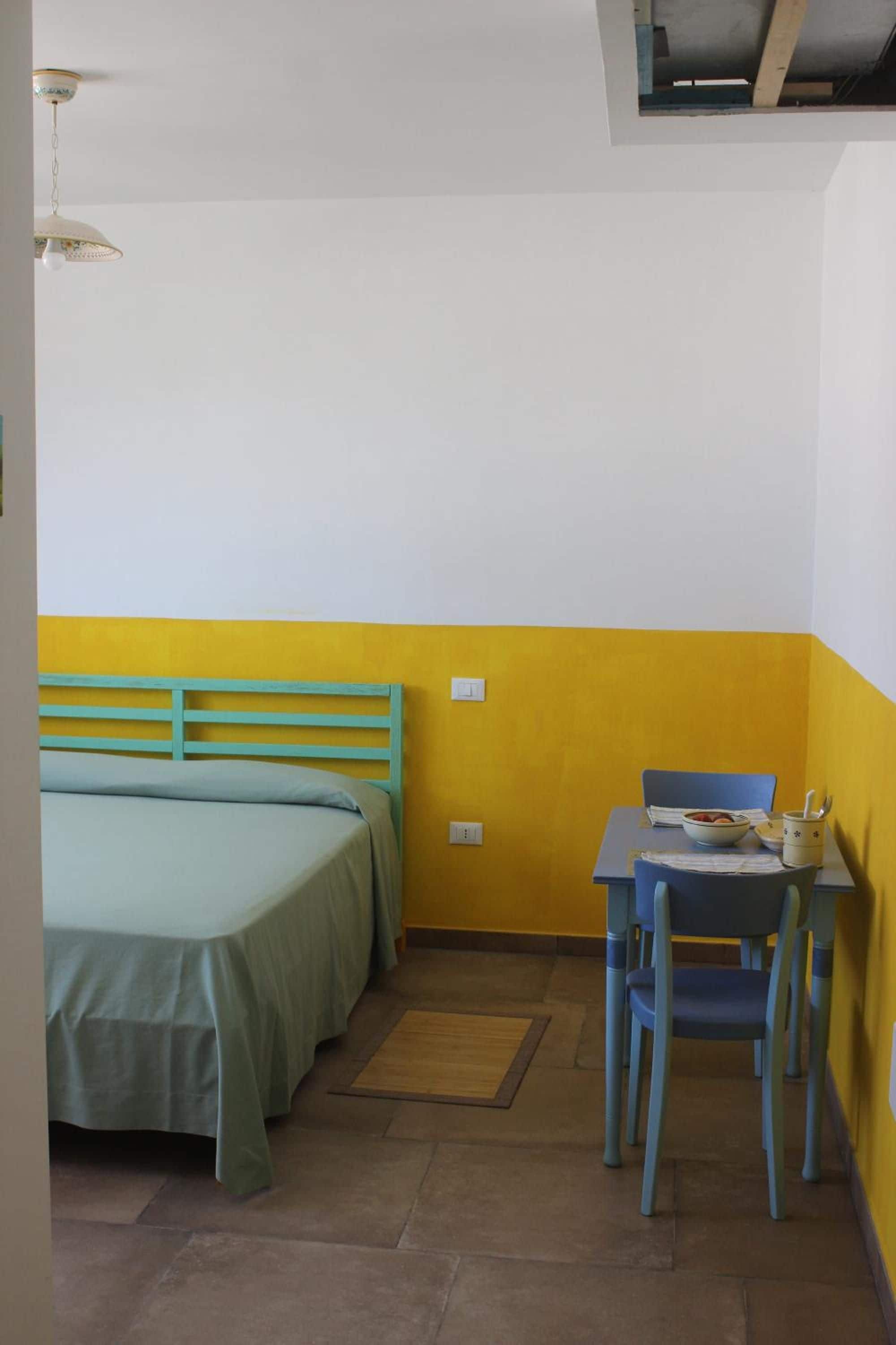 Quarto pequeno e compacto, com cama, mesa e cadeiras, além de uma janela.