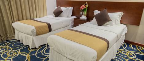Quarto de hotel com duas camas, uma cabeceira de madeira, mesa de cabeceira com um vaso de flores e carpete estampado.