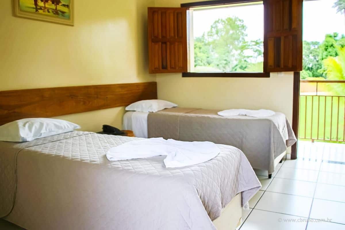 Quarto de hotel com duas camas, cabeceira de madeira, janela com vista para vegetação e varanda com guarda-corpo.