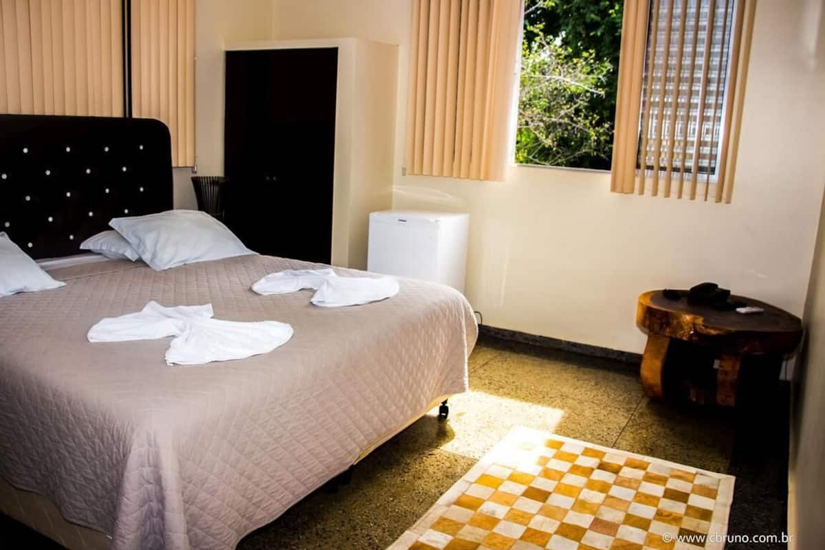 Quarto de hotel com cama, mesinha, cadeira, radiador e janela com persianas.
