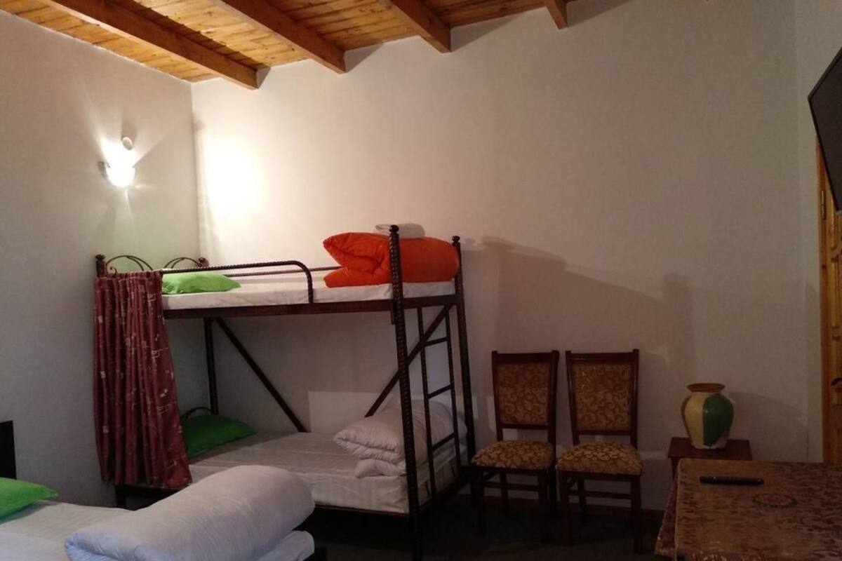 Um quarto com beliche, duas cadeiras, uma mesinha e um ponto de luz fixado na parede.