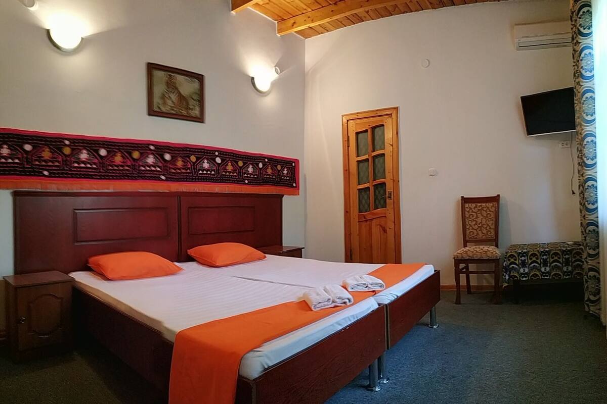 Quarto com cama de madeira, duas mesas de cabeceira, porta de madeira, uma cadeira e uma televisão na parede.