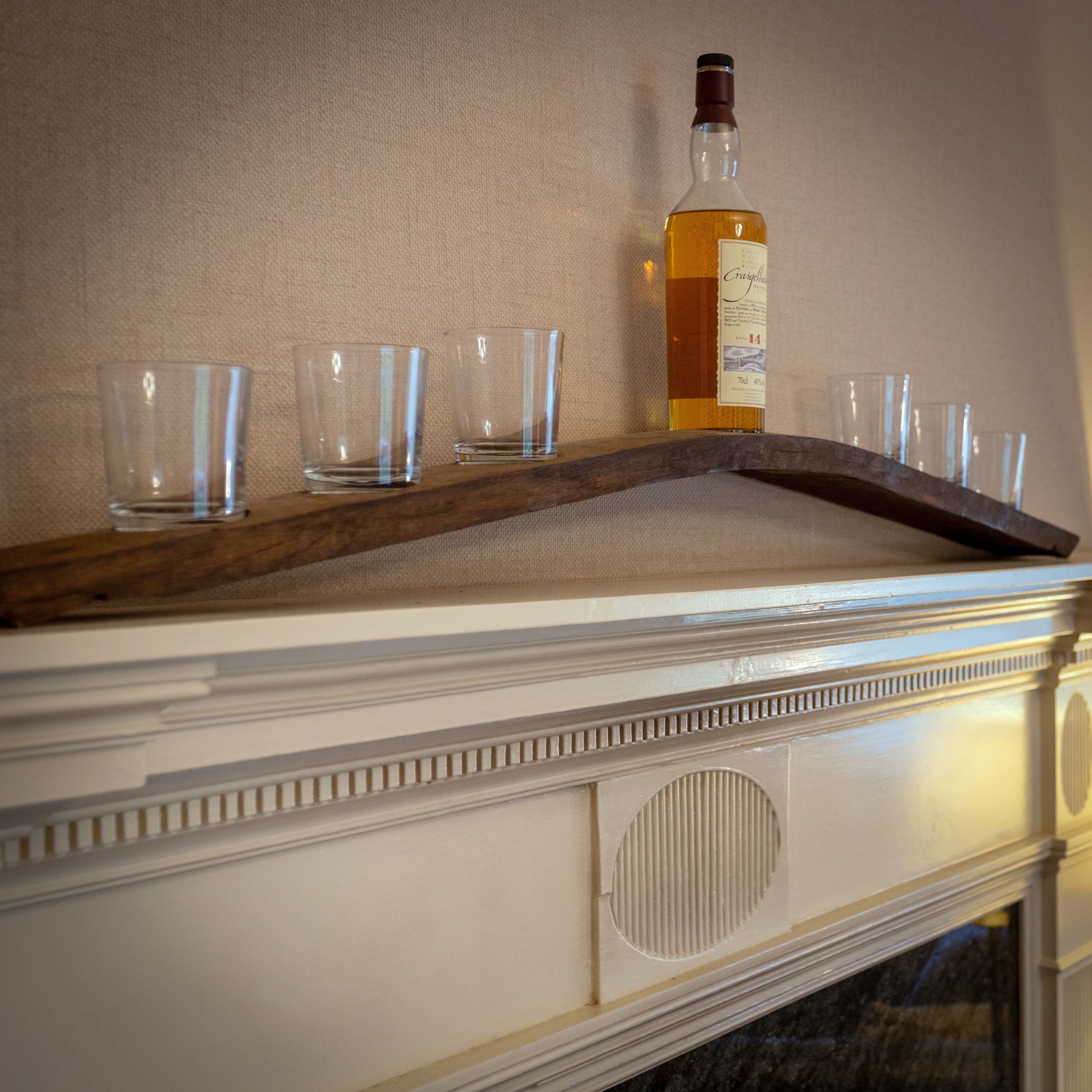 Uma garrafa de bebida alcoólica sobre uma bandeja de madeira, com vários copos em uma lareira de cor branca.
