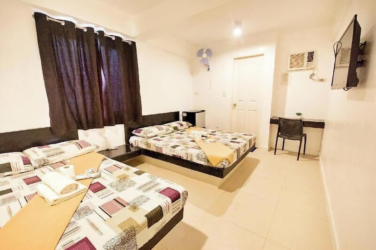 Quarto com duas camas, uma escrivaninha, uma cadeira, um ventilador e uma porta.