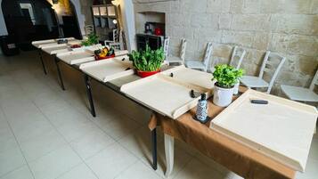 Uma mesa comprida com toalha marrom, cadeiras brancas e plantas em vasos.