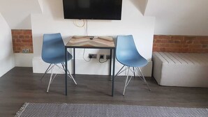 Um espaço de estar pequeno e moderno, com televisão na parede, uma mesa pequena, duas cadeiras azuis, um sofá e um tapete estampado.