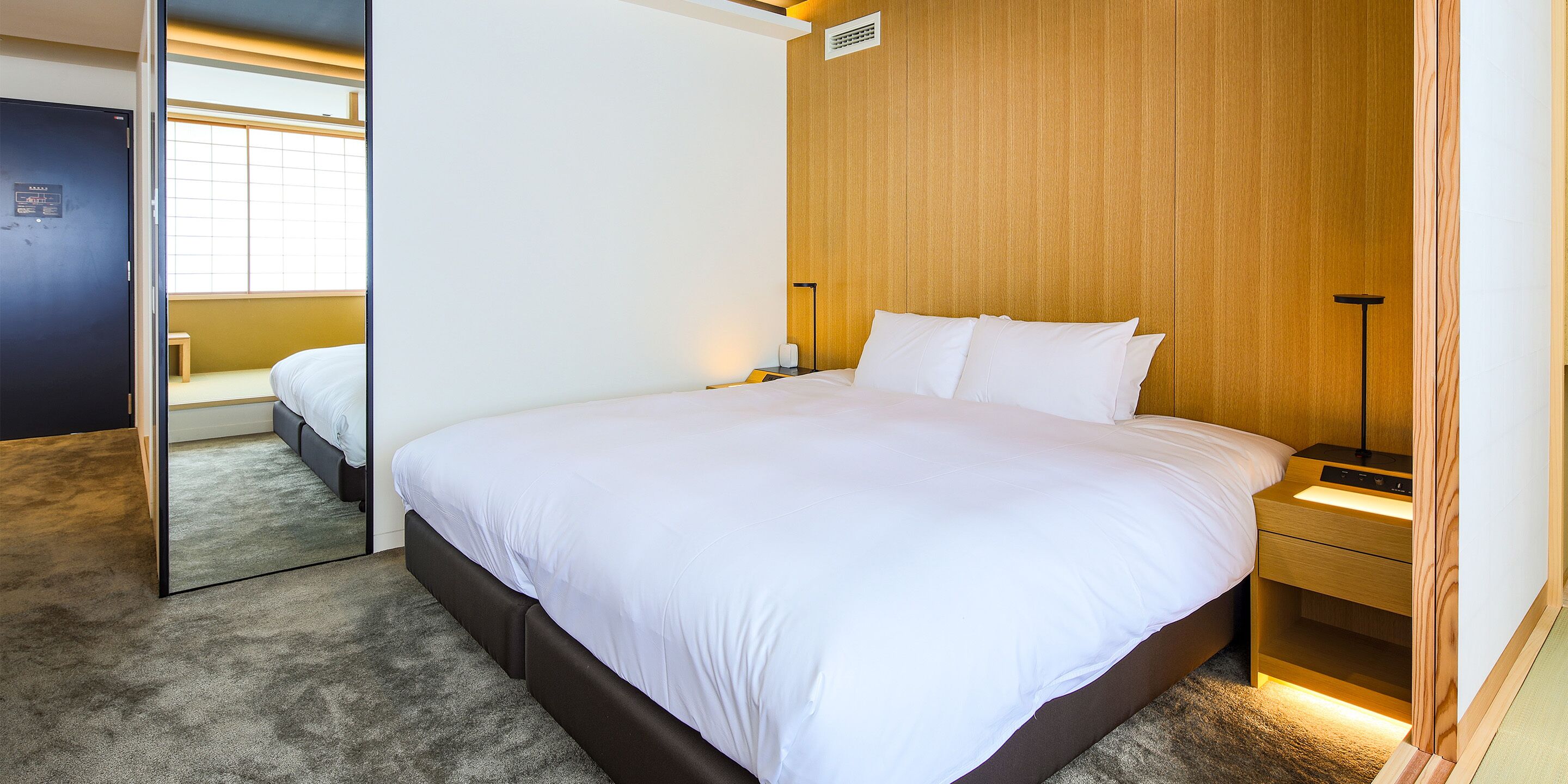 Quarto de hotel moderno com uma cama grande, mesa de cabeceira com luminária e cabeceira de madeira.
