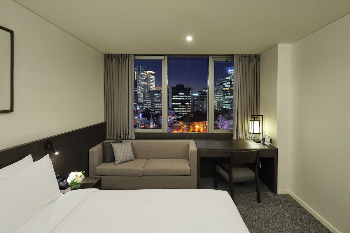 Quarto de hotel com cama, sofá, escrivaninha e vista para a cidade à noite.