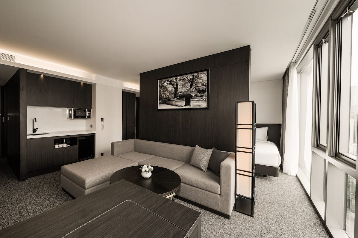 Quarto de hotel moderno com sofá, mesa de centro, cama, pequena cozinha e uma janela grande.