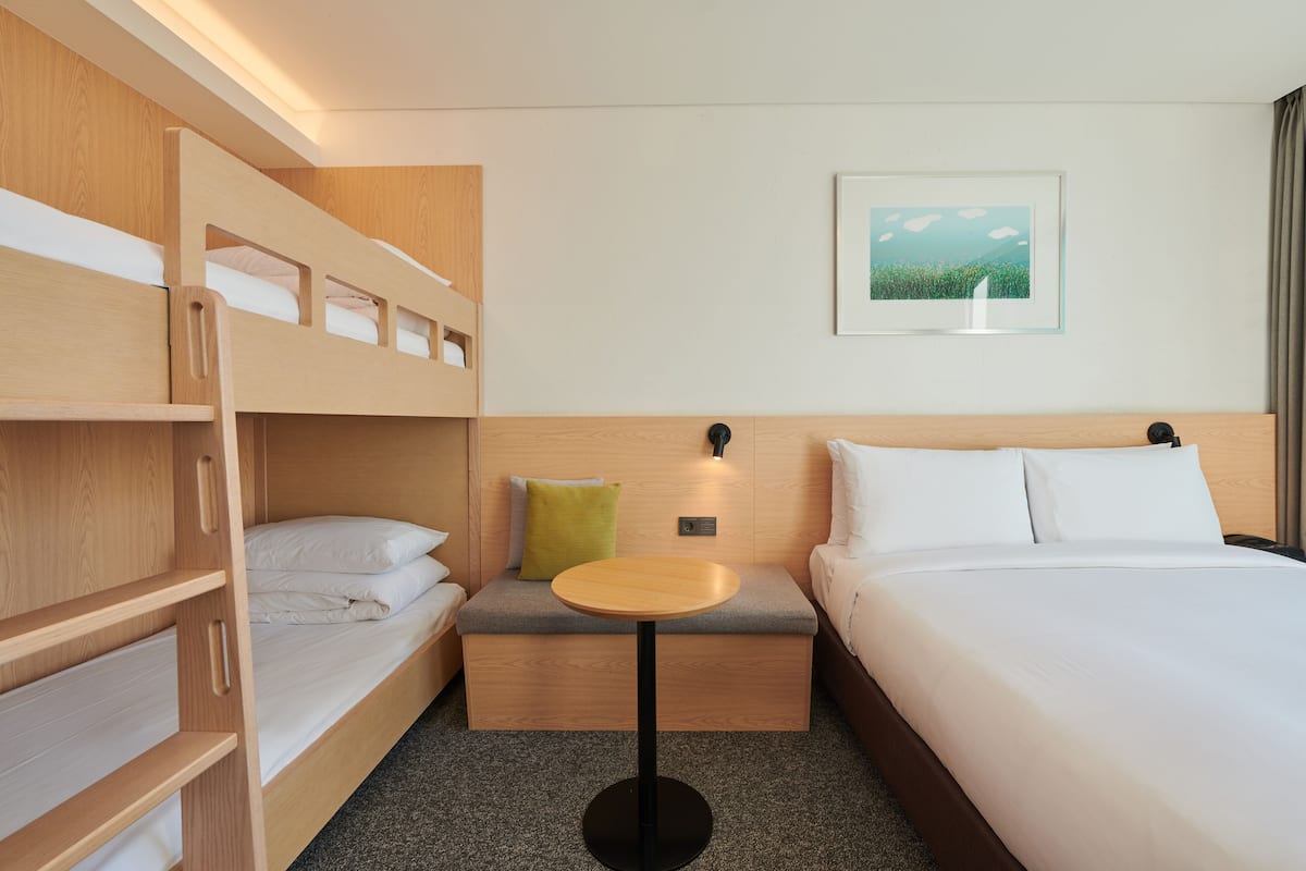 Quarto de hotel com beliche, uma cama de solteiro, uma mesa pequena e um quadro na parede.