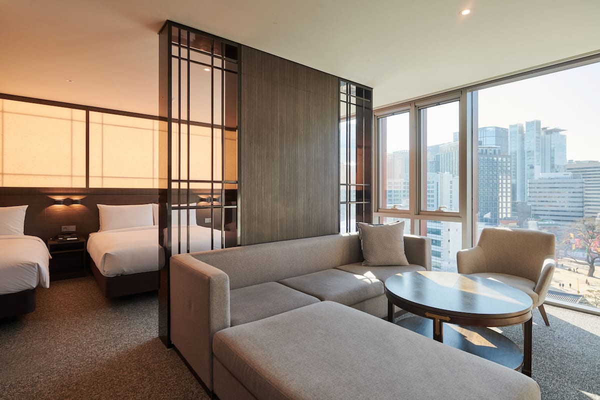 Quarto de hotel moderno com uma cama grande, um sofá, uma mesa pequena e vista para o horizonte da cidade.