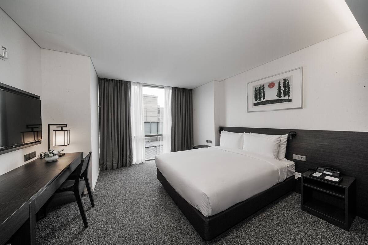 Quarto de hotel com uma cama, uma escrivaninha, uma cadeira, uma televisão, uma mesa de cabeceira e um quadro pendurado na parede.