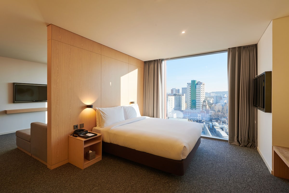 Quarto de hotel moderno com uma cama grande, mesa de cabeceira com abajur, televisão na parede e vista da cidade através de uma janela ampla.