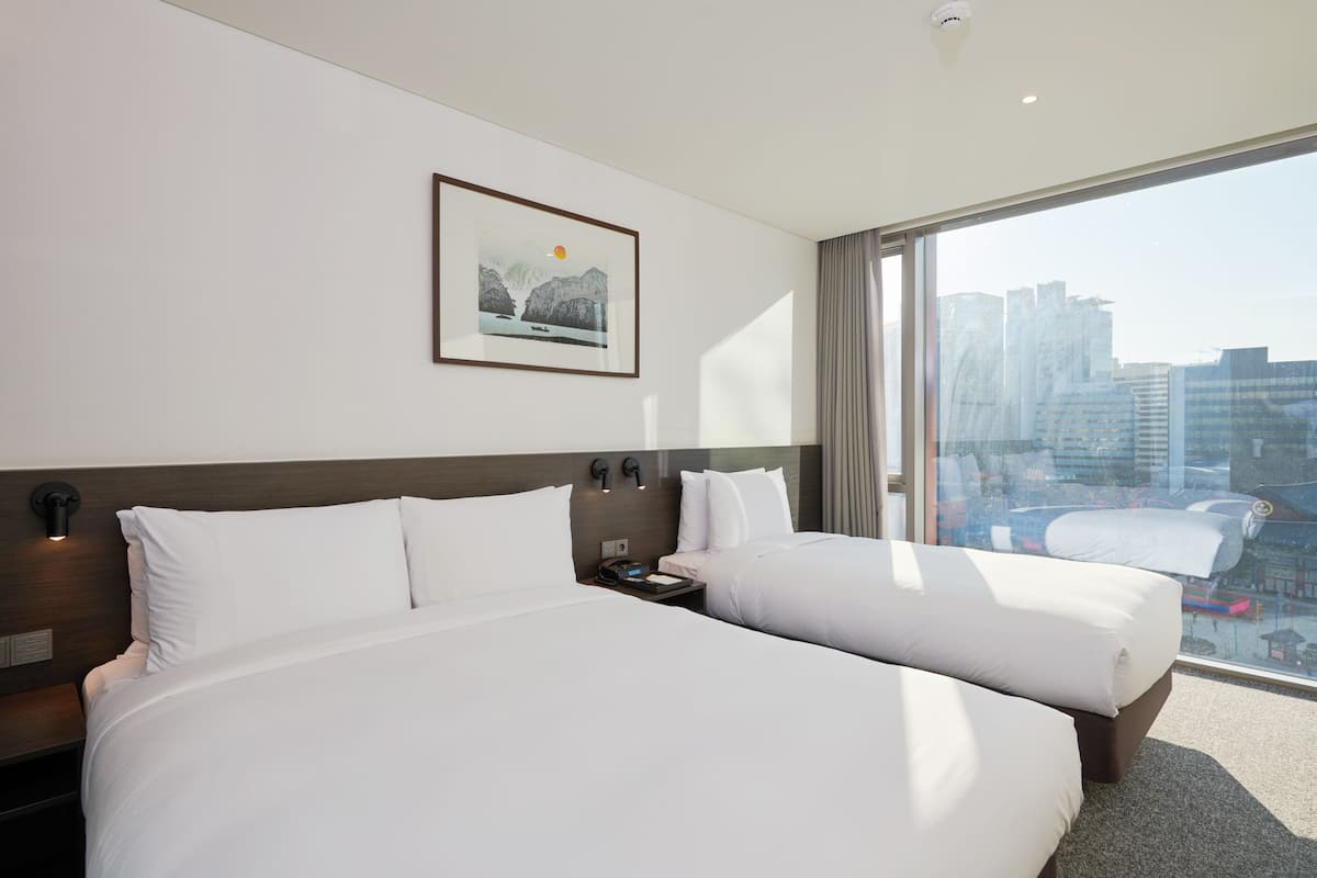 Quarto de hotel com duas camas, uma mesa de cabeceira, luminária fixa na parede, quadro emoldurado e uma janela grande com vista para a cidade.