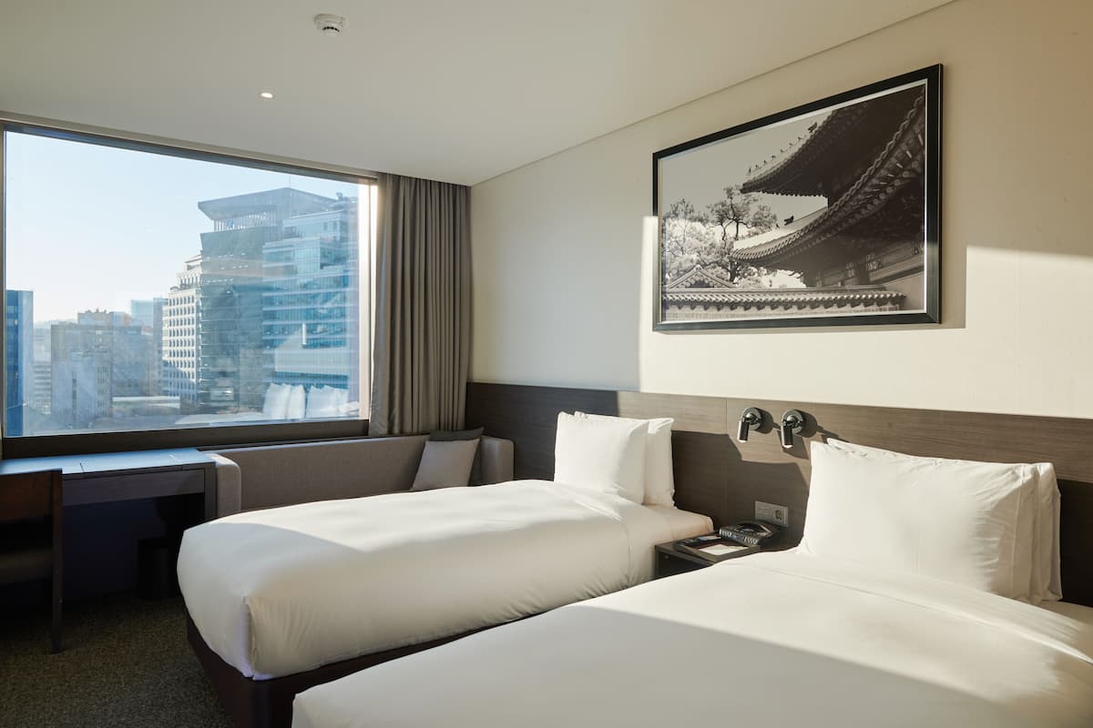 Quarto de hotel com duas camas, uma escrivaninha e uma janela grande com vista para a cidade.
