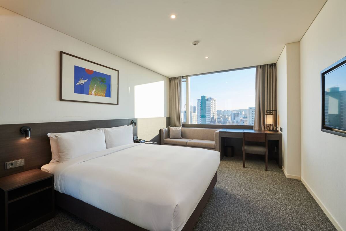 Quarto de hotel com uma cama grande, uma escrivaninha, um sofá e vista para a cidade.