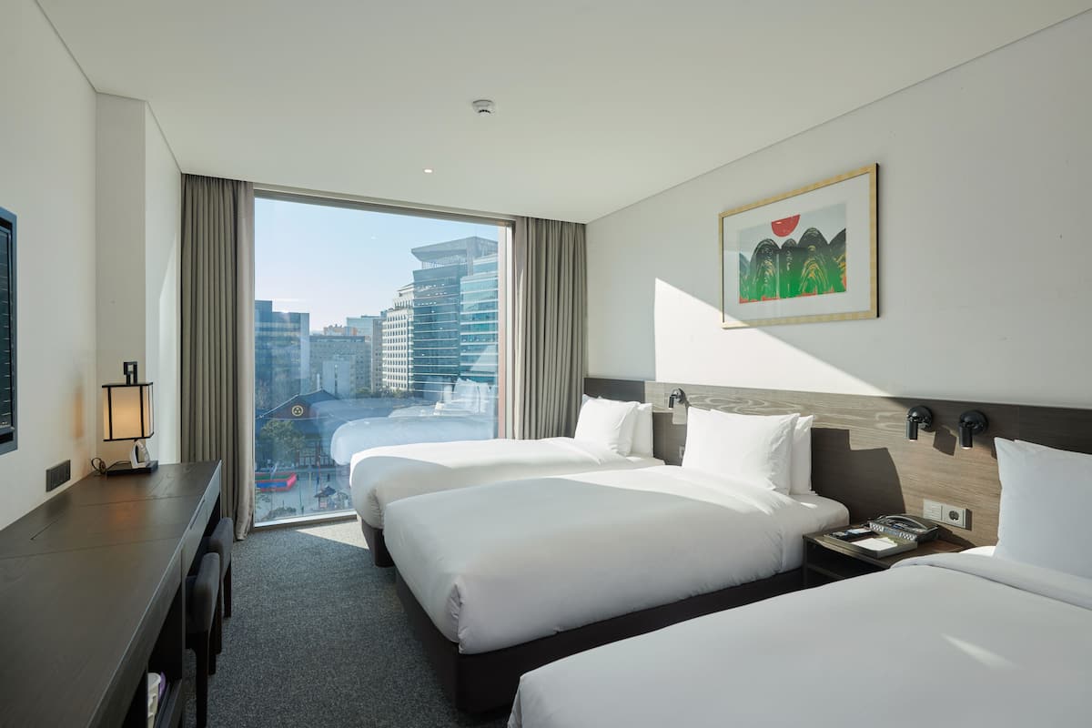 Quarto de hotel com duas camas, uma escrivaninha, uma luminária e uma janela grande com vista para a cidade.