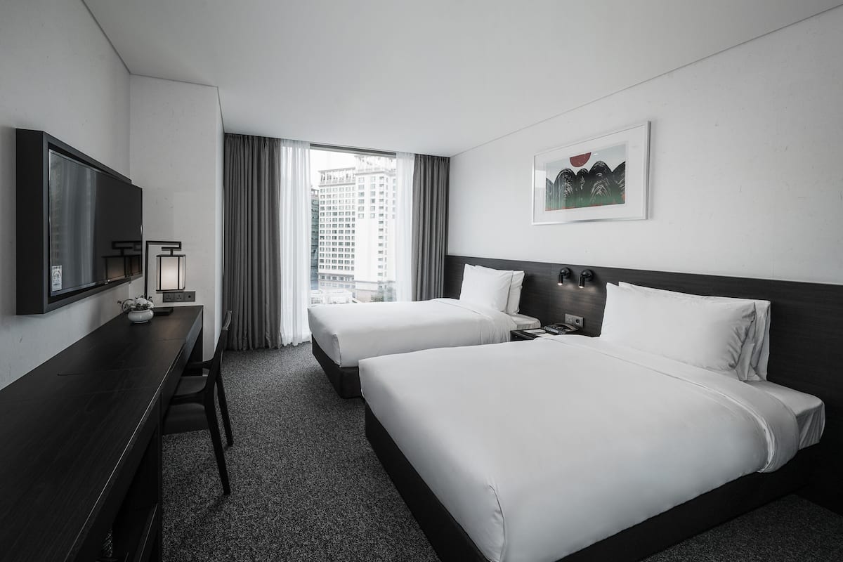 Quarto de hotel com duas camas, uma escrivaninha, televisão e janela vista para a cidade.