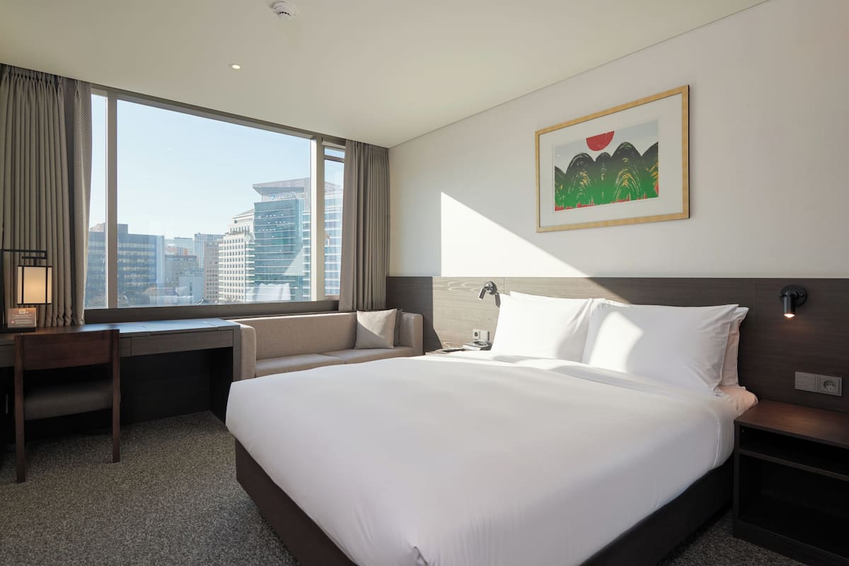 Quarto de hotel com uma cama grande, uma escrivaninha, uma cadeira e uma janela com vista para a cidade.