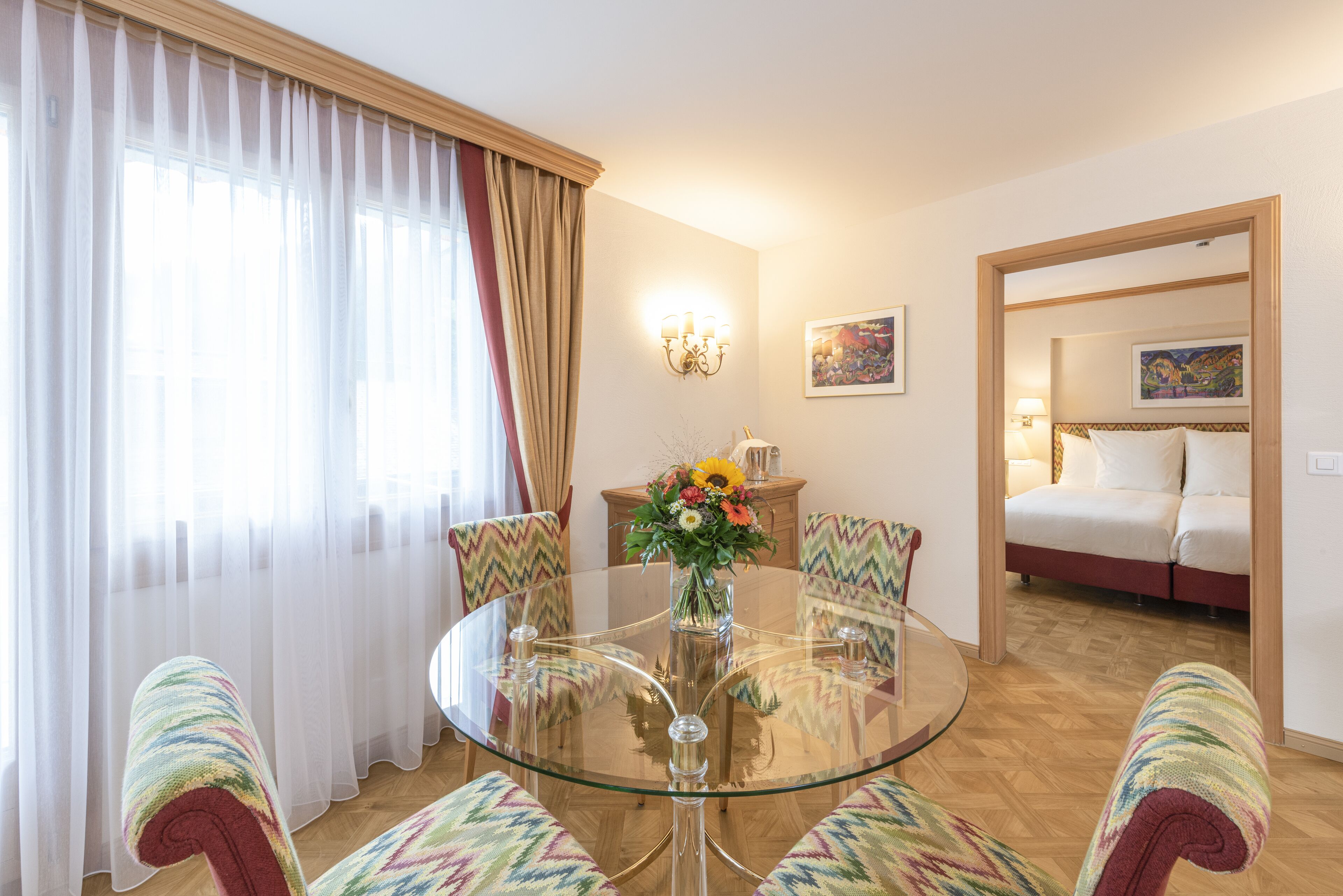 Quarto de hotel com uma mesa de vidro redonda, quatro cadeiras estampadas, uma cama, uma mesinha de cabeceira com abajur e um quadro na parede.
