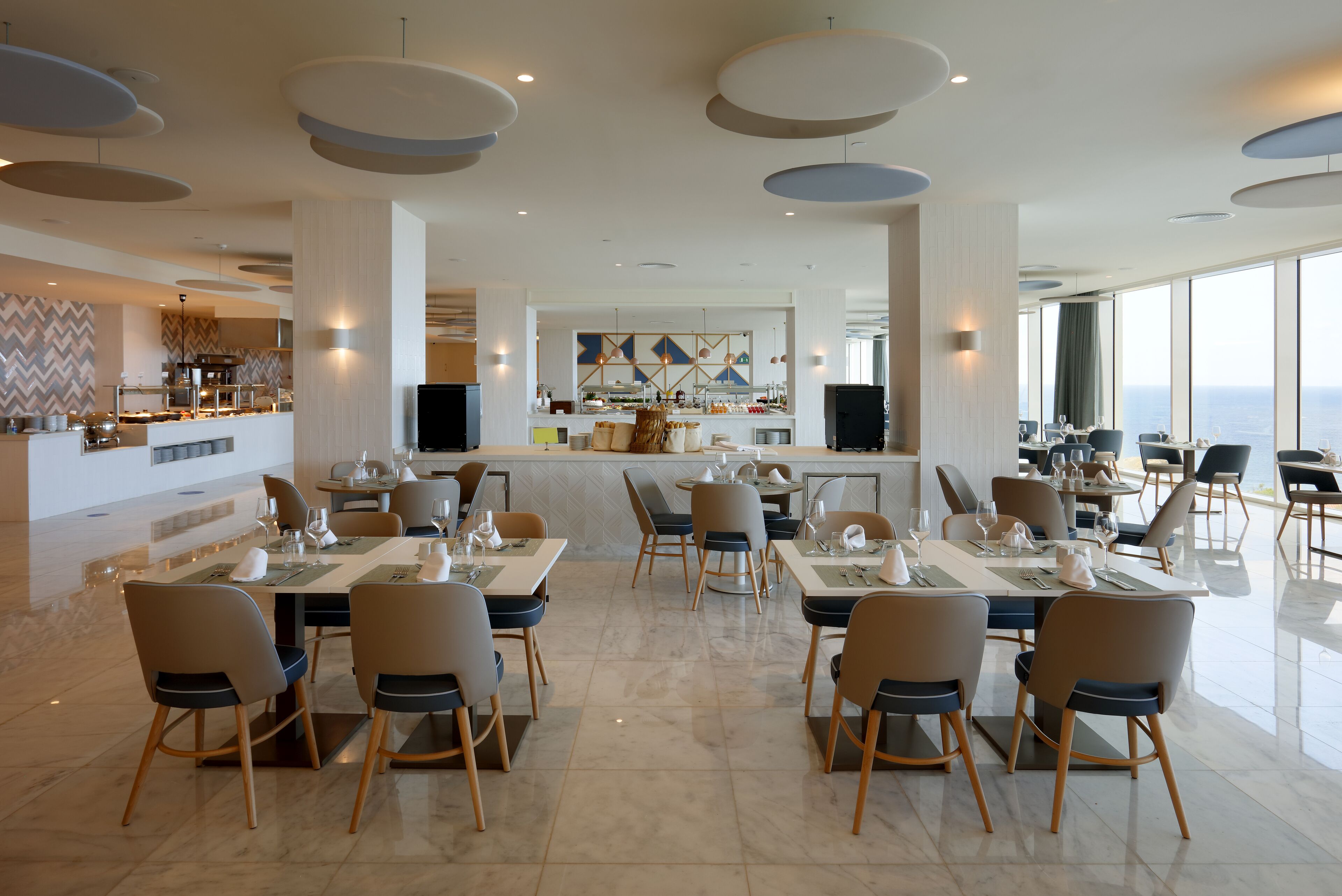 Restaurante moderno com várias mesas e cadeiras, um balcão central e grandes janelas com vista para o oceano.