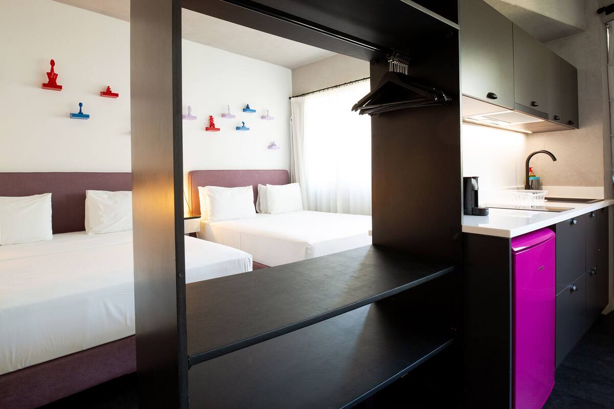Quarto de hotel com duas camas, uma pequena cozinha e um espelho refletindo o espaço.