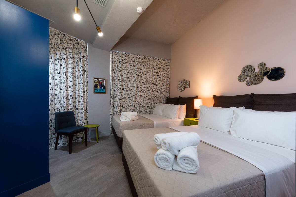 Quarto de hotel com duas camas, uma cadeira e uma mesa pequena.