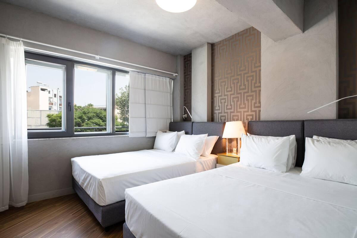 Quarto de hotel com duas camas, uma janela com cortinas, parede estampada e mesa de cabeceira com abajur.