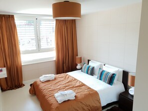 Quarto de hotel com uma cama grande, duas luminárias de cabeceira, uma janela com cortinas e vista para prédios.